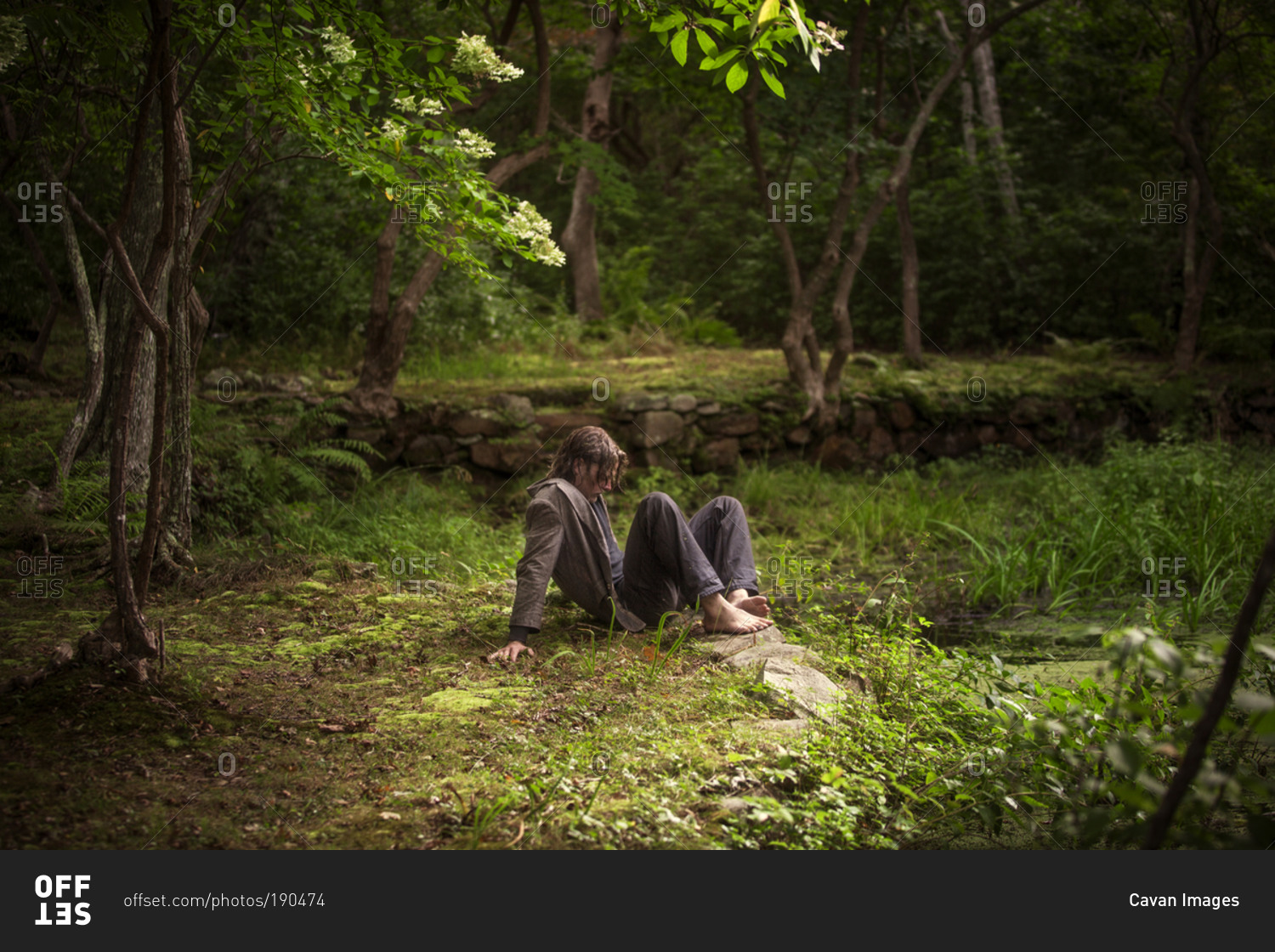 Young man sitting on ground in woods thinking stock photo - OFFSET