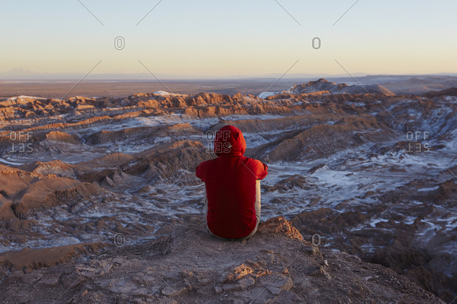 Man in a hoodie watching a sunset from a cliff in a desert valley ...