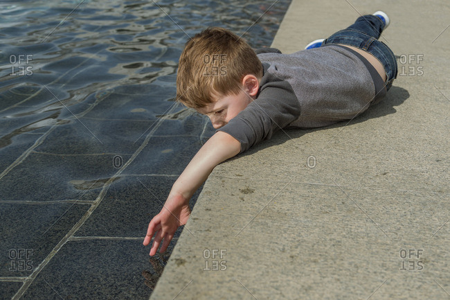 Boy lying on a sidewalk putting his hand in a shallow fountain pool ...