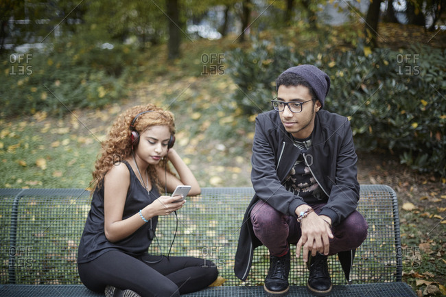 Young couple with cell phone and headphones on park bench - Stock Image ...