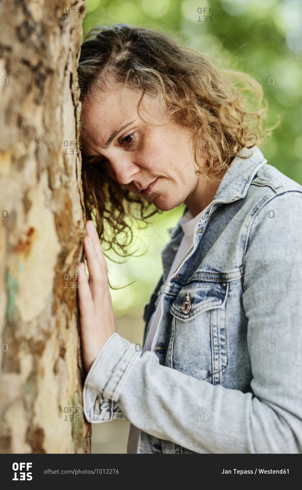 Close-up of thoughtful woman wearing denim jacket leaning on tree trunk ...