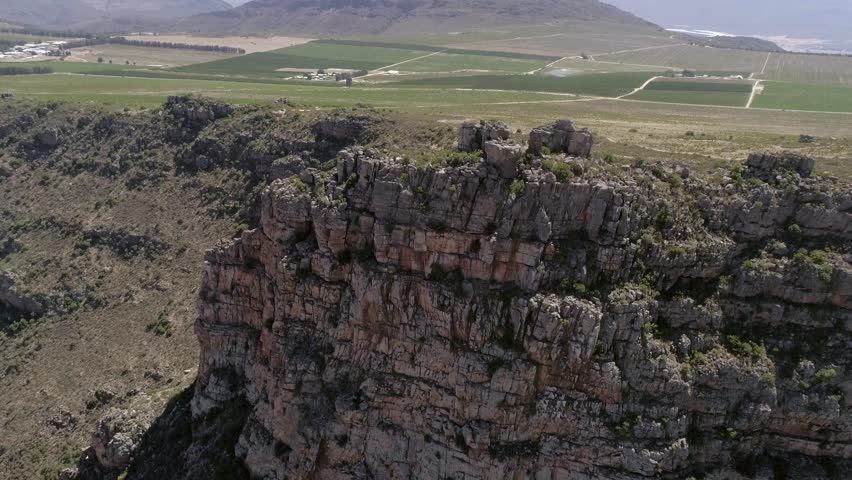 Aerial descends rugged rock escarpment, vineyards, orchards beyond ...